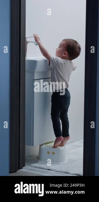 Baby boy stands on toes to open water tap. Kid is happy to watch the ...