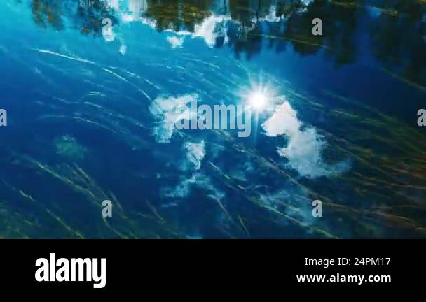 Sun is shining through the surface of a body of water reflecting the blue sky and white clouds ...