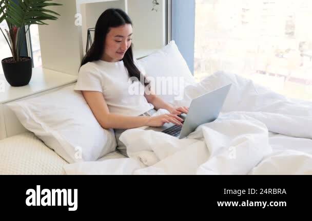 Asian woman in her 20s using laptop in cozy bedroom setting. Relaxed ...