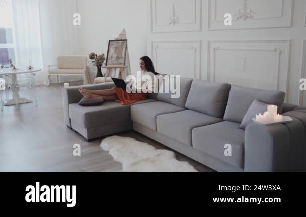 A woman is seated comfortably on a studio couch in a living room, using ...