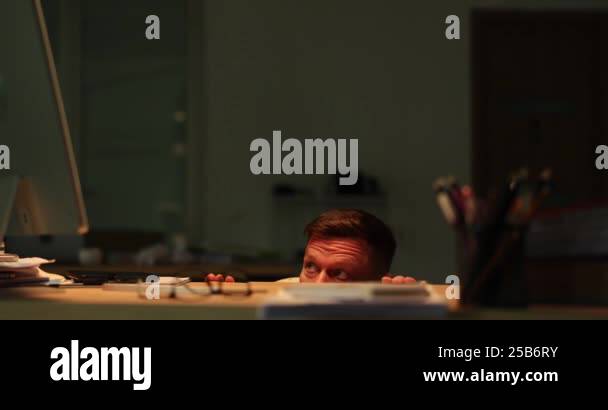 Frightened woman hides under desk glancing around in dark office ...