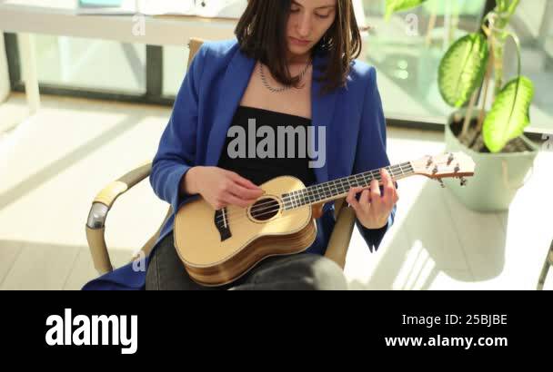 Young woman plays ukulele strumming strings with concentration. Brunette female in blazer ...