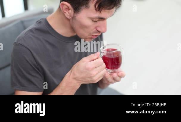 Man in casual T-shirt sips tea sitting on sofa in living room. Guy with ...