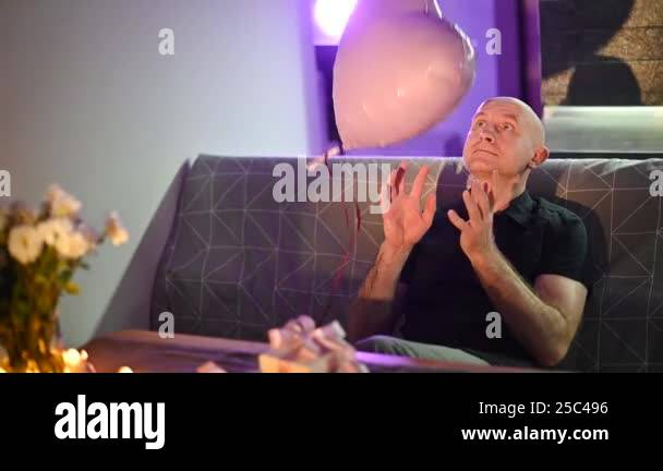 Bald man is catching a heart shaped balloon while sitting on a couch in ...