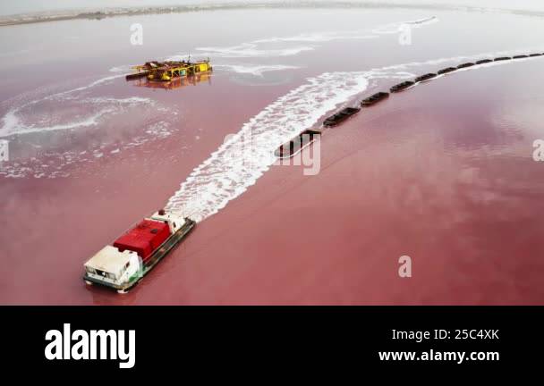 Motorboat pulls cargo barges near mining platform on pink salt ...