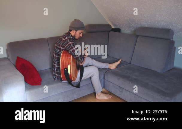 Musician sits indoors on a cozy couch, strumming an acoustic guitar in ...