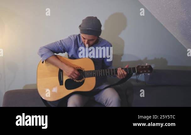 Musician sits indoors on a cozy couch, strumming an acoustic guitar in ...