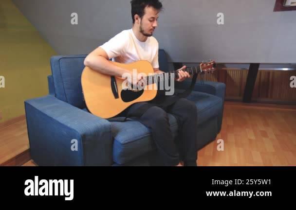 Musician sits indoors on a cozy couch, strumming an acoustic guitar in ...
