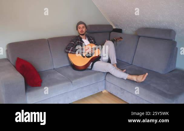 Musician sits indoors on a cozy couch, strumming an acoustic guitar in ...
