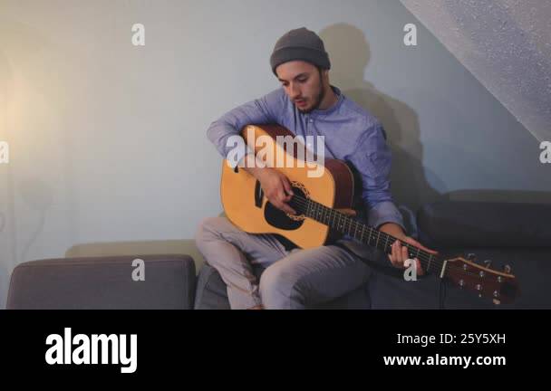 Musician sits indoors on a cozy couch, strumming an acoustic guitar in ...