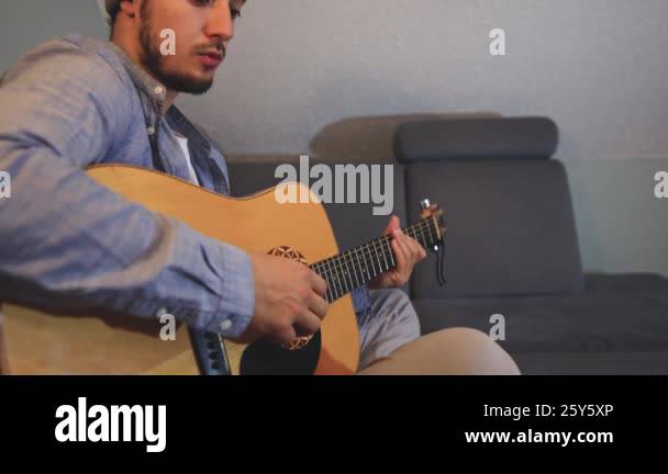 Musician sits indoors on a cozy couch, strumming an acoustic guitar in ...
