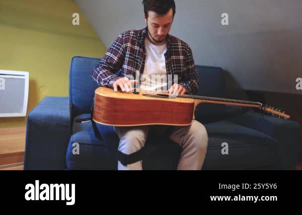 Musician sits indoors on a cozy couch, strumming an acoustic guitar in ...