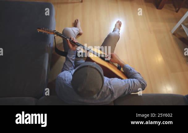 Musician sits indoors on a cozy couch, strumming an acoustic guitar in ...