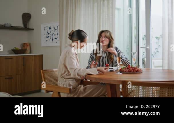 Smiling ladies talking at kitchen table looking fashion magazine. Two ...