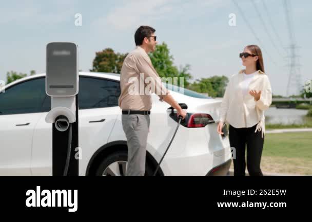 Slow motion couple using smartphone to pay EV car battery at charging station connected to power ...