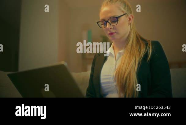 A professional woman in a suit works remotely from home on her laptop late at night, focusing on ...