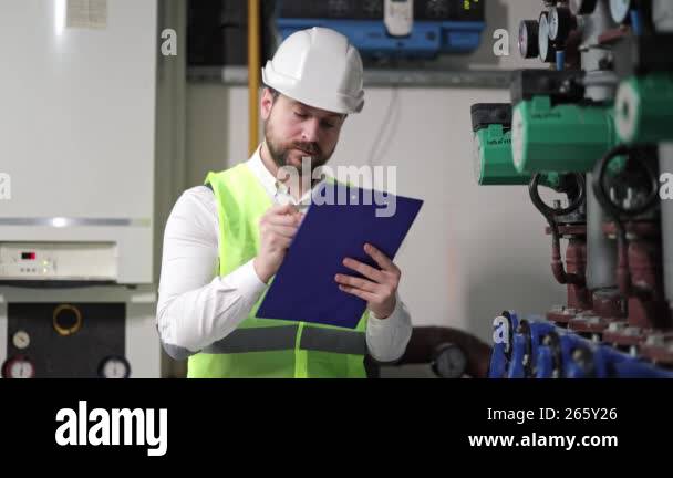 Caucasian male boiler room engineer in safety vest and helmet writes on ...