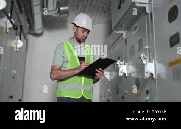 Serious electrical engineer in a hard hat and uniform inspects an ...