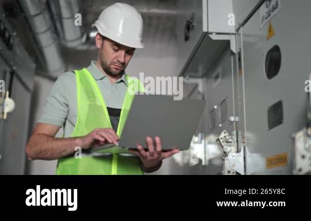 Serious electrical engineer in a uniform and a helmet types on a laptop ...