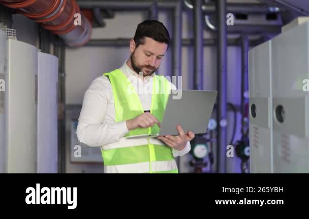 Technician wearing safety vest uses laptop in boiler room. Industrial environment with pipelines ...