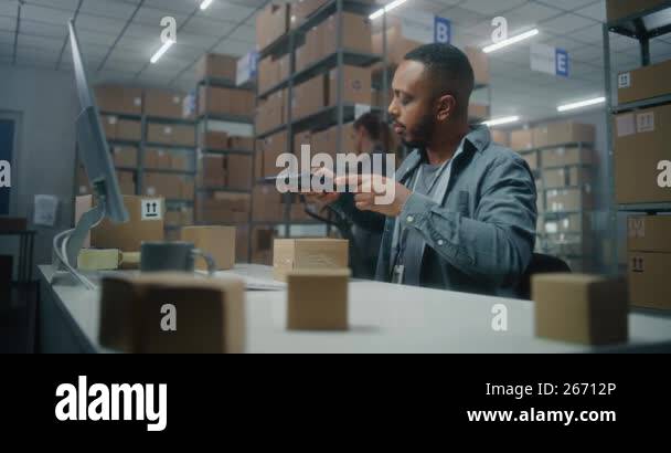 African American warehouse associate scans parcel using tablet computer, looks at PC screen ...