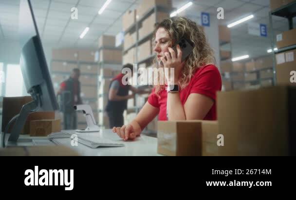 Female customer support specialist in logistics talks to customer on mobile phone, uses computer ...