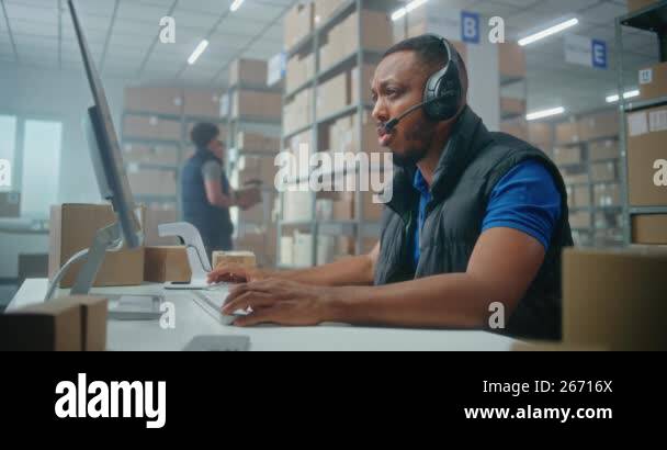 African American logistics customer support specialist in headset talks to client, uses computer ...
