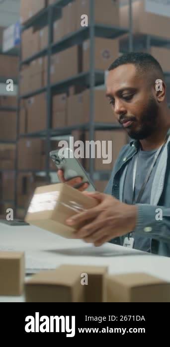African American logistics manager scans parcel using smartphone, works in post office warehouse ...