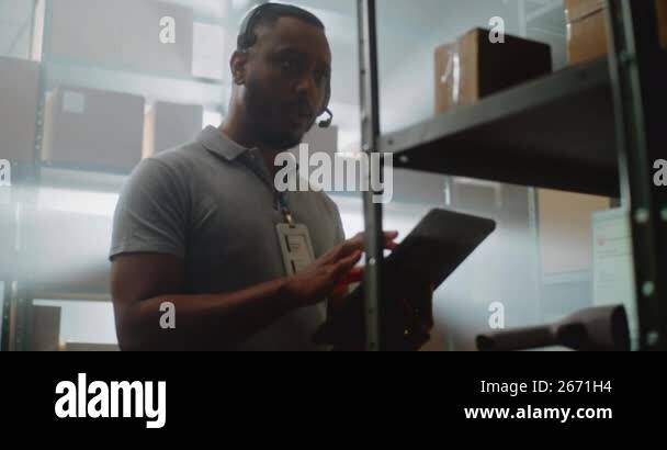 African American Warehouse Associate Talks on Headset, Checks Information Using Tablet Computer ...