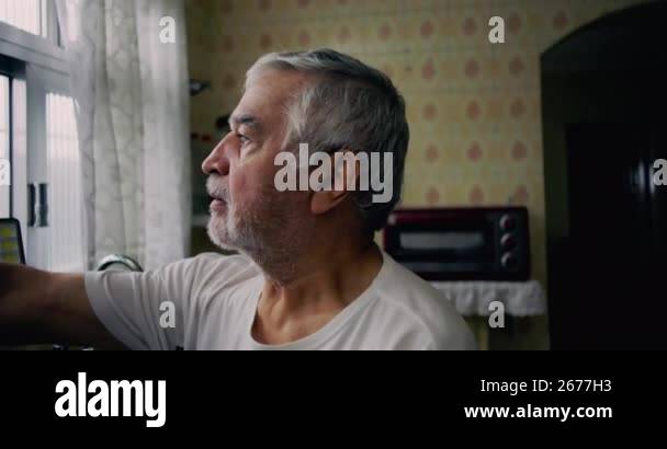 Elderly man gazing out of kitchen window with introspective expression ...