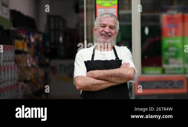 Elderly male shopkeeper wearing an apron smiling with arms crossed ...