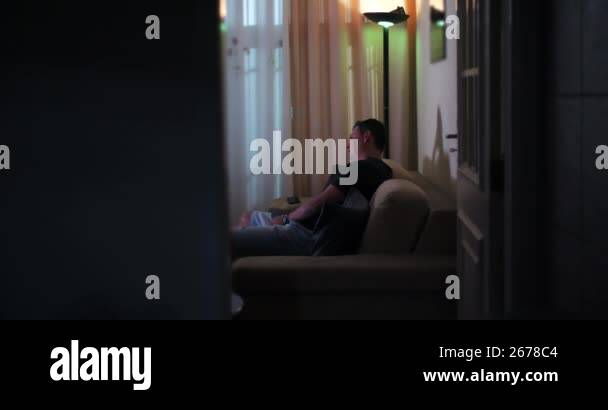 Side view of a man leaning back on a couch, TV remote in hand, watching ...