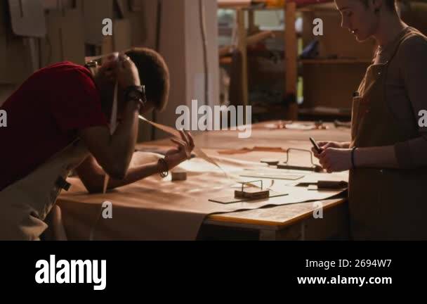 Zoom-in shot of young concentrated multiethnic artisans measuring leather details on workbench ...