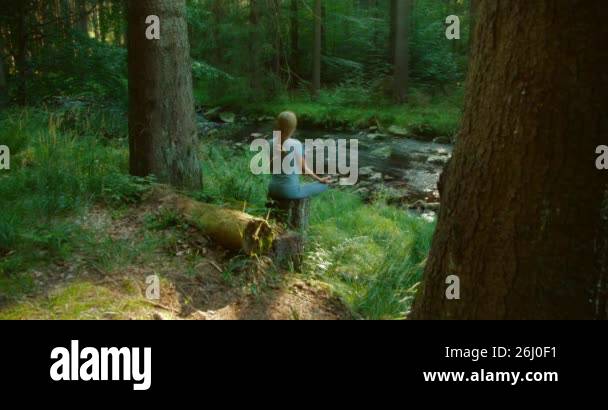 Woman sits in yoga pose on a tree stump in the woods near a flowing ...