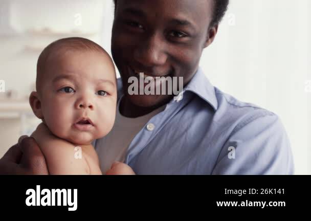 In a cozy home setting, a father smiles while holding his baby close ...