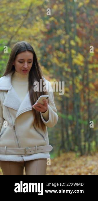 Walking brunette focused on her phone in autumn park. Long-haired lady ...