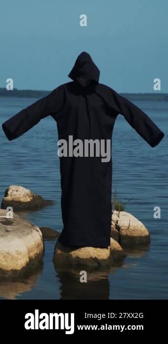 Woman devil in black cape and hood on stones near the river. Halloween ...