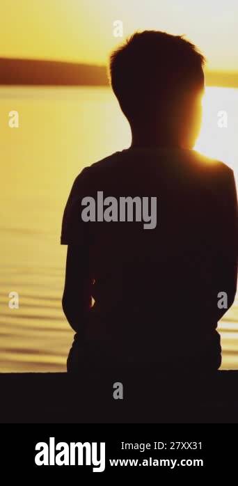 Little boy sitting on wooden dock at sunset. Child is watching the ...