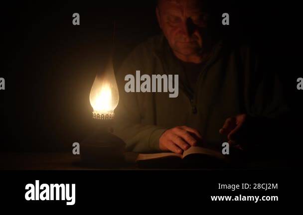 Man reads a book by the light of a kerosene lamp at night time, close ...
