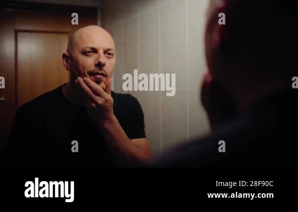 A bald man in a black t-shirt gazes at his reflection in a bathroom ...