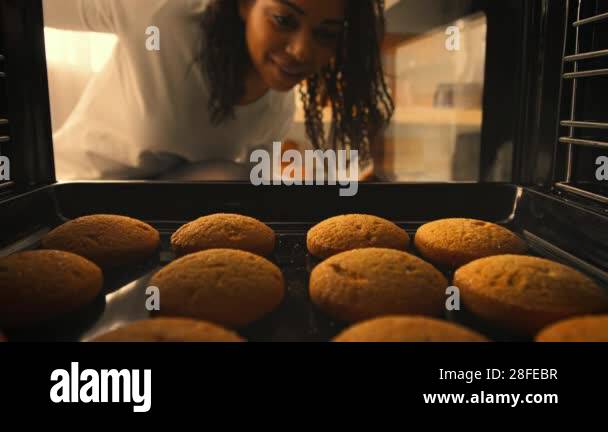 A millennial woman smiles as she peers into an oven filled with freshly ...