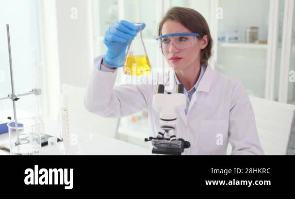 Scientist leans closer to beaker to observe yellow liquid. Concentrated woman chemist checks ...