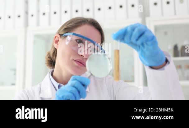 Chemist uses magnifying glass to study properties of liquid. Woman lab assistant searches for ...