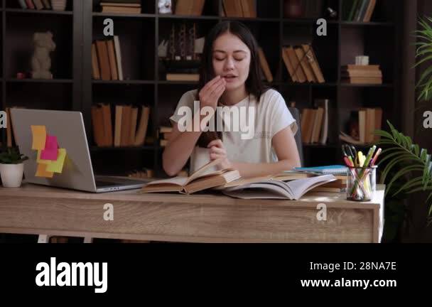 Tired young woman yawns with hand over mouth studying for exams in a ...