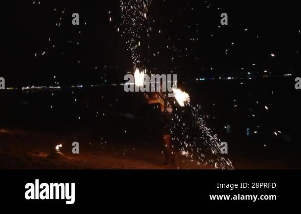 A performer manipulates fire props on a beach at night, with sparks and ...