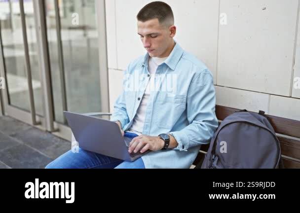 A focused man working on a laptop while seated on a bench with a backpack beside him in a modern ...