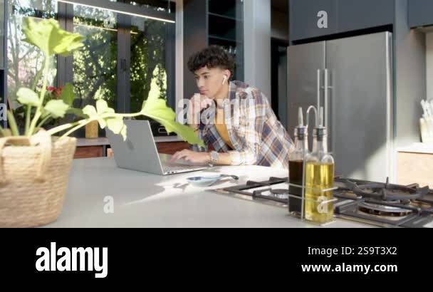 Using laptop in kitchen, teenage boy with earbuds focusing on work ...