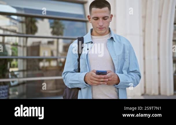 A young man with a backpack texting on his phone in front of a modern ...