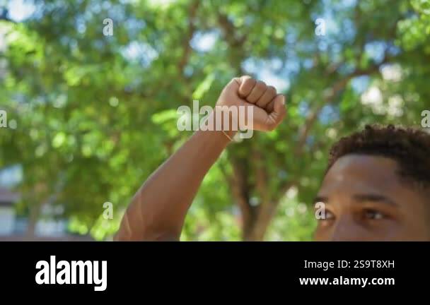Young man raising fist in urban park setting with trees behind ...