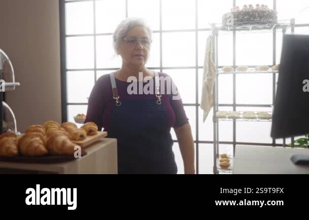 Elderly woman wearing apron standing behind bakery counter with ...